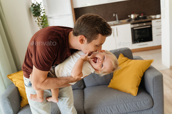Father cuddling, hugging, tickling baby son after returning home from work holding him in hands ...
